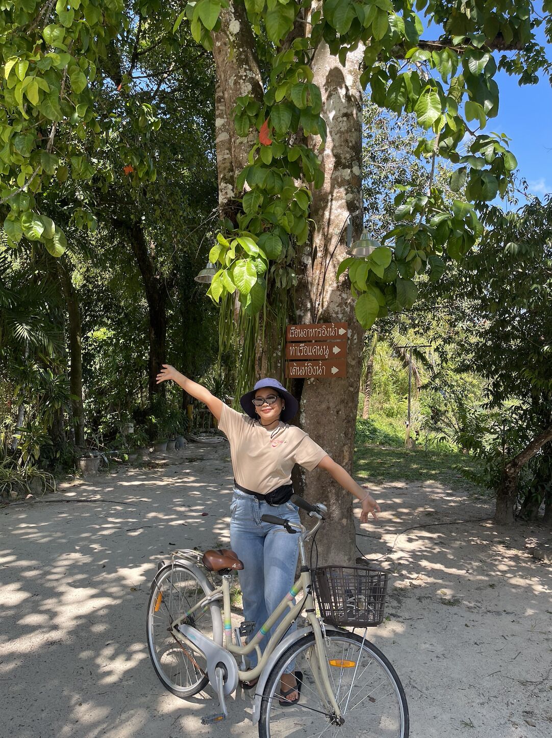 Uma pessoa ao lado de uma bicicleta com cesta, em ambiente natural com árvores e uma placa presa a um tronco.