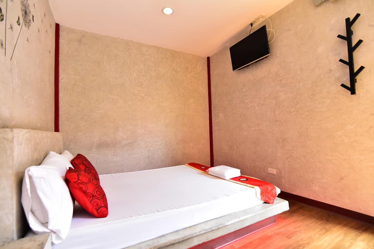 Uma cama bem arrumada com roupa de cama branca e vermelha, uma televisão de tela plana na parede e um chão de madeira.