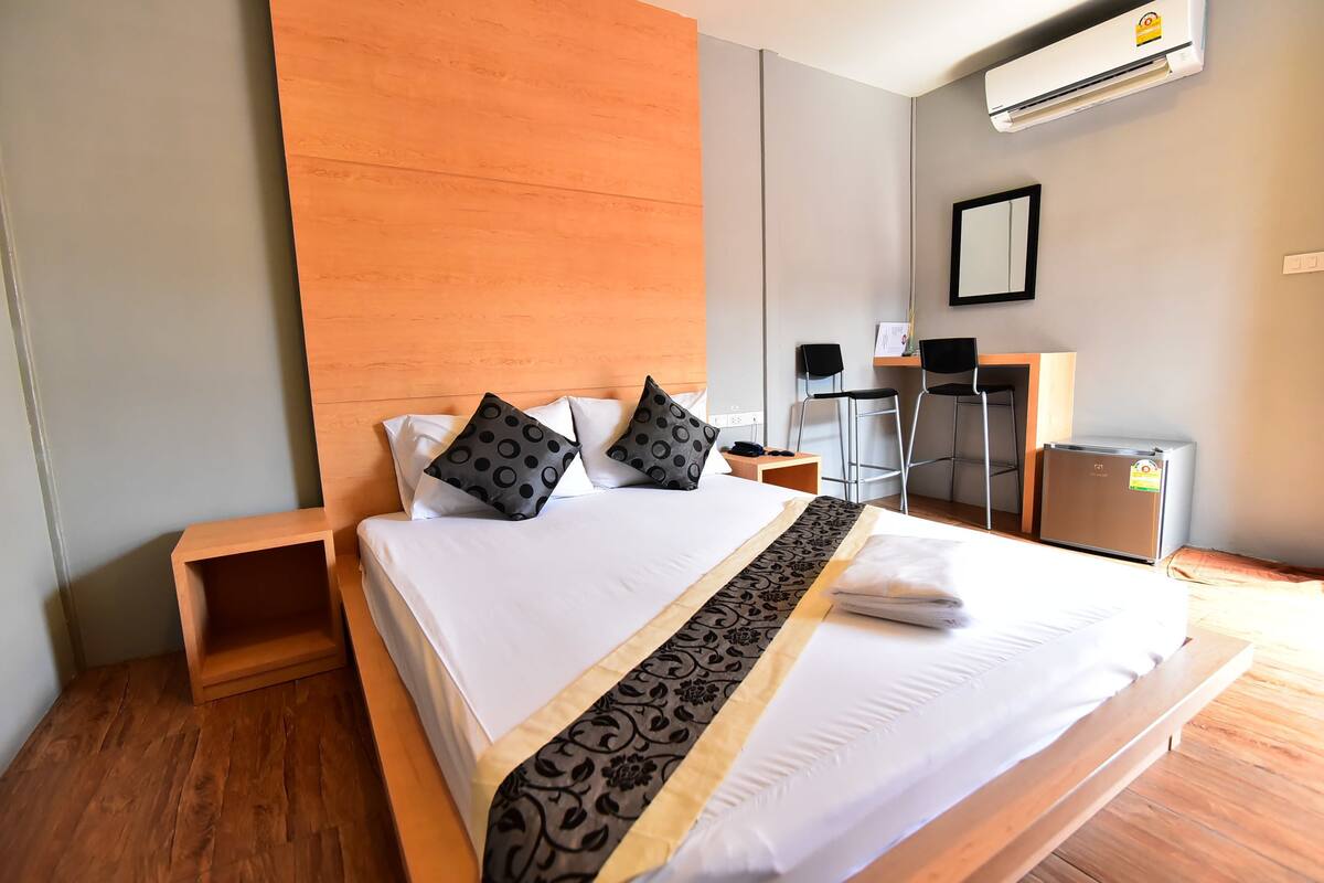 Quarto de hotel com uma cama grande, cabeceira de madeira, escrivaninha com cadeira e pequeno frigorífico.