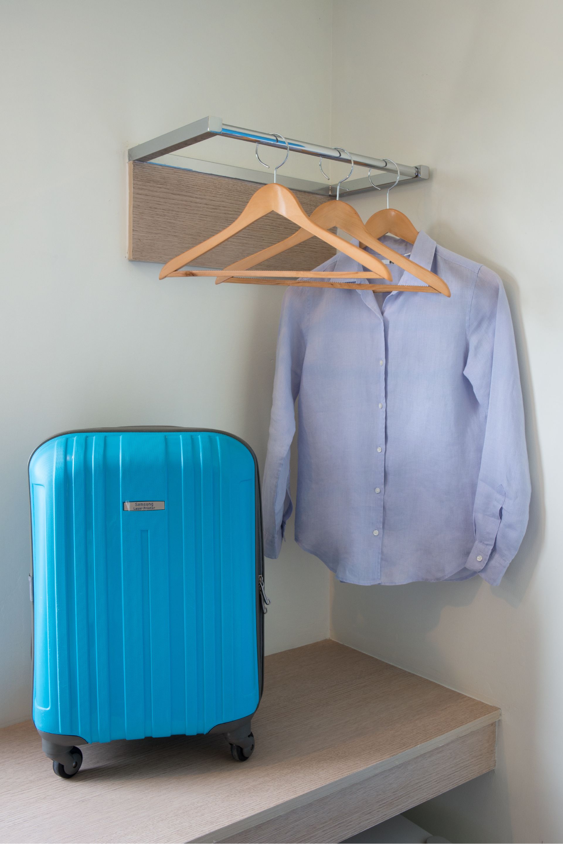 Quarto com mala azul sobre um banco de madeira, um cabideiro de madeira com roupas e uma camisa azul clara pendurada.