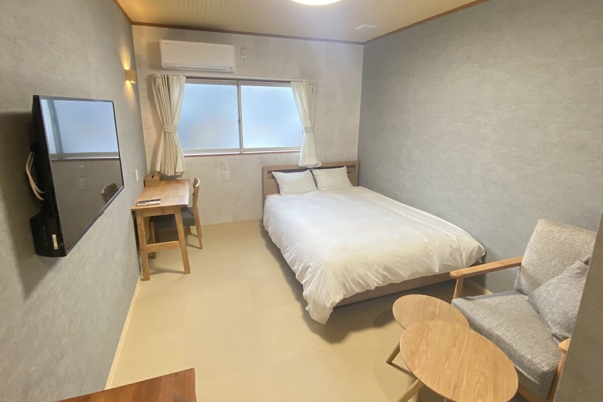 Quarto pequeno e moderno com uma cama, uma escrivaninha, uma cadeira e uma televisão.