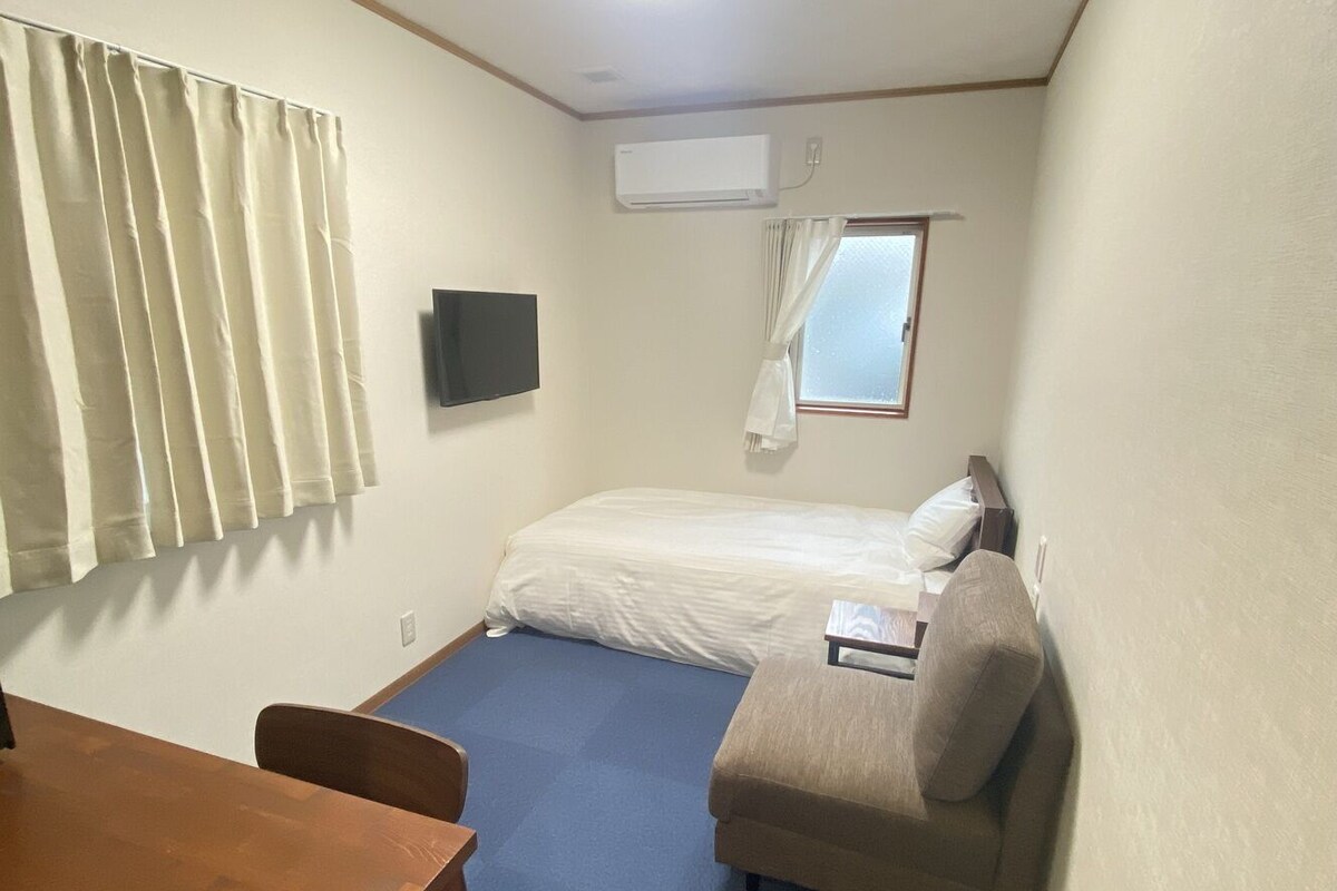 Acomodação compacta, em um único ambiente, com cama, sofá, mesa e ar condicionado.