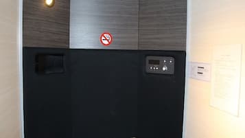 Quarto de canto com uma cama, um painel de controle, um aviso de proibido fumar e um interruptor de luz.