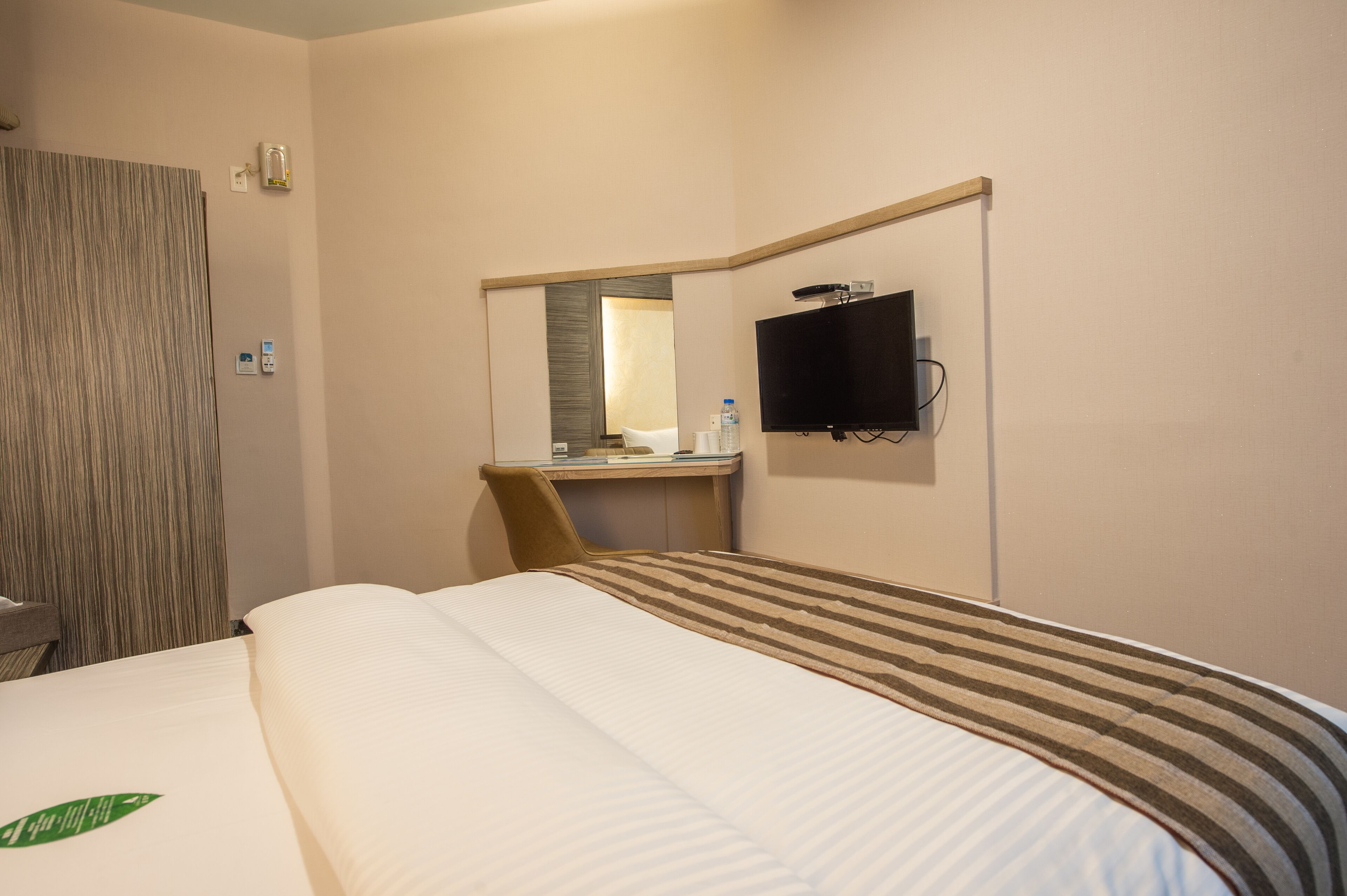 Quarto de hotel com cama, escrivaninha com cadeira, TV de tela plana na parede e espelho sobre a escrivaninha.