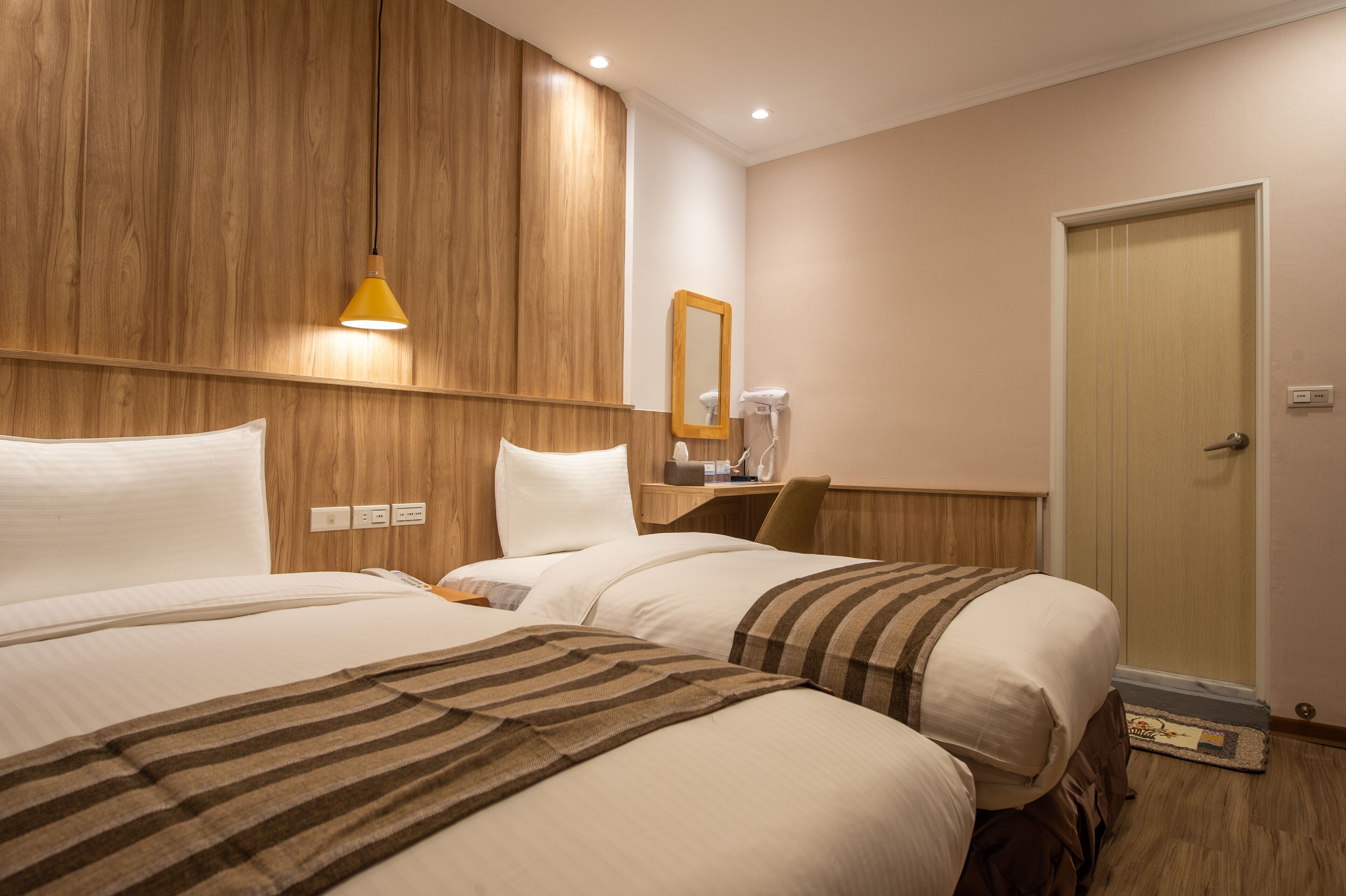 Quarto de hotel com duas camas, painéis de madeira, um espelho, uma escrivaninha e uma porta.