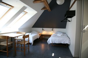 Quarto pequeno e bem iluminado, com teto inclinado, duas camas, uma mesa e cadeiras.