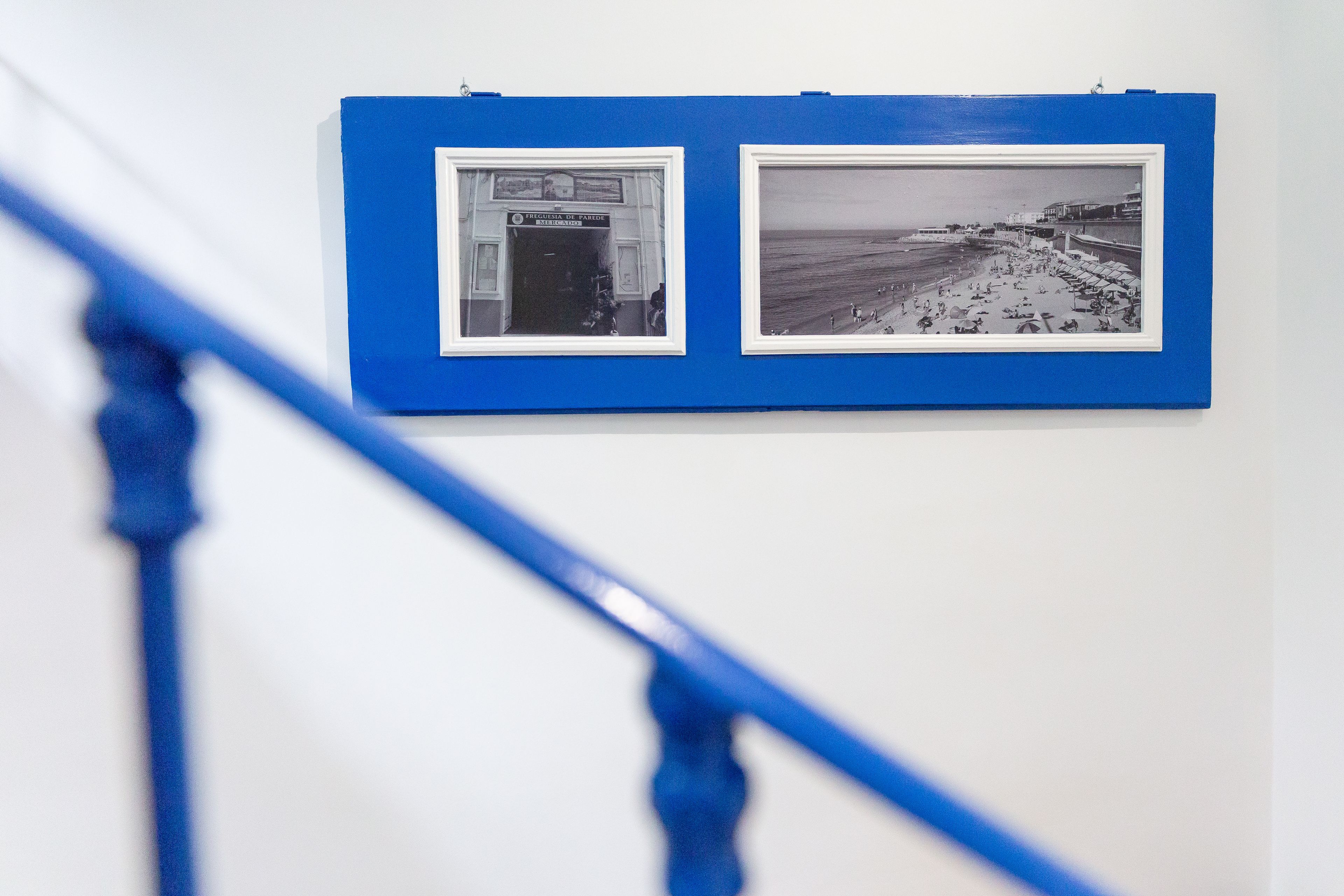 Uma escadaria com corrimão azul e duas fotografias emolduradas na parede.