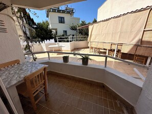 Uma varanda com mesa e cadeiras, plantas em vasos e vista para edifícios vizinhos.