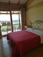 Quarto com cama grande, teto de madeira, varanda com vista para o verde e uma cadeira.