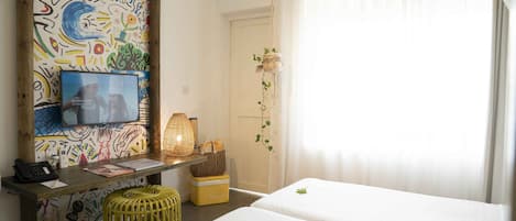 Quarto de hotel com duas camas, uma secretária com telefone e papéis, uma parede colorida com arte abstrata e uma janela com cortinas leves.