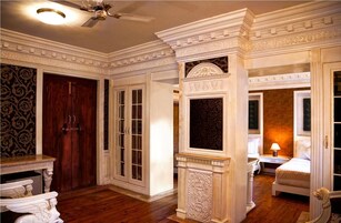 Quarto com piso de madeira, uma grande porta de madeira, parede branca com um padrão decorativo, lareira branca e uma cama com colcha branca e dourada, além de uma mesa de cabeceira com abajur.