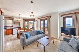 Uma sala de estar moderna com sofá, mesa de centro e televisão. Há uma área de cozinha com frigorífico, pia e mesa de jantar. O espaço possui grandes janelas e vista para o exterior.