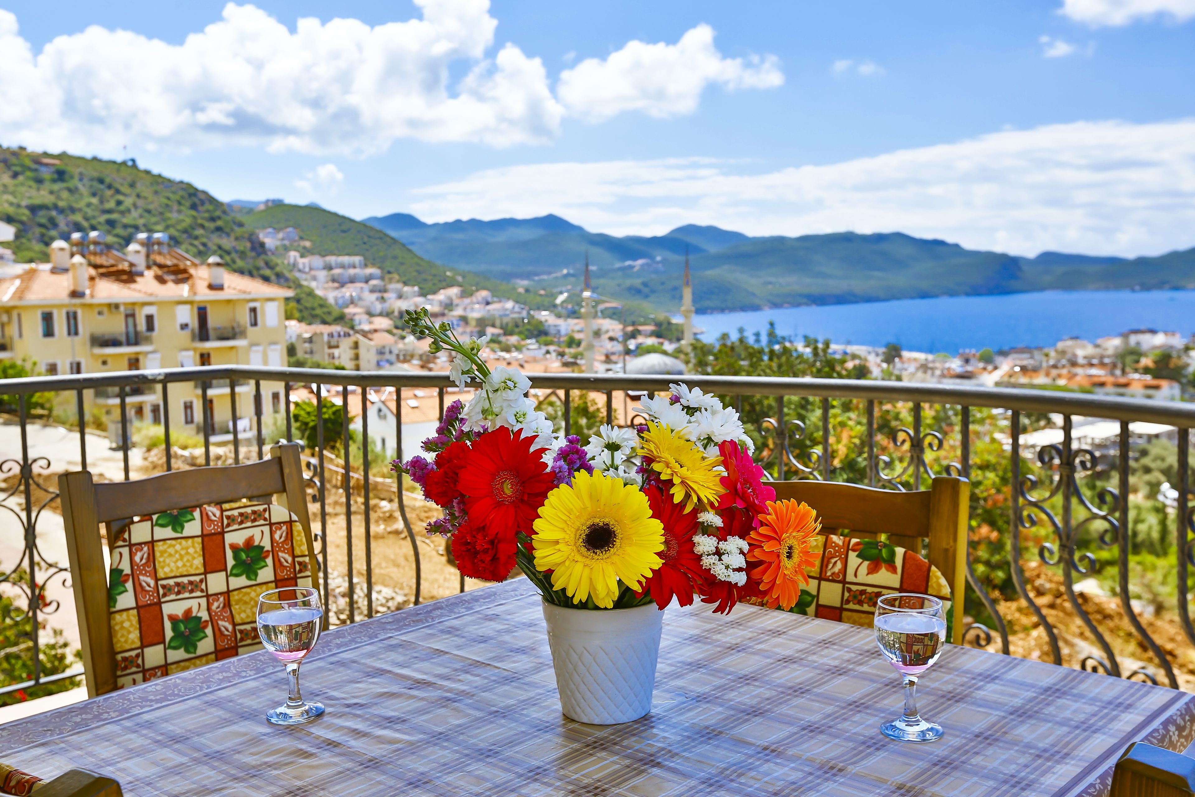 Uma varanda com mesa posta para dois, um vaso de flores coloridas e uma vista panorâmica de uma cidade e montanhas ao fundo.