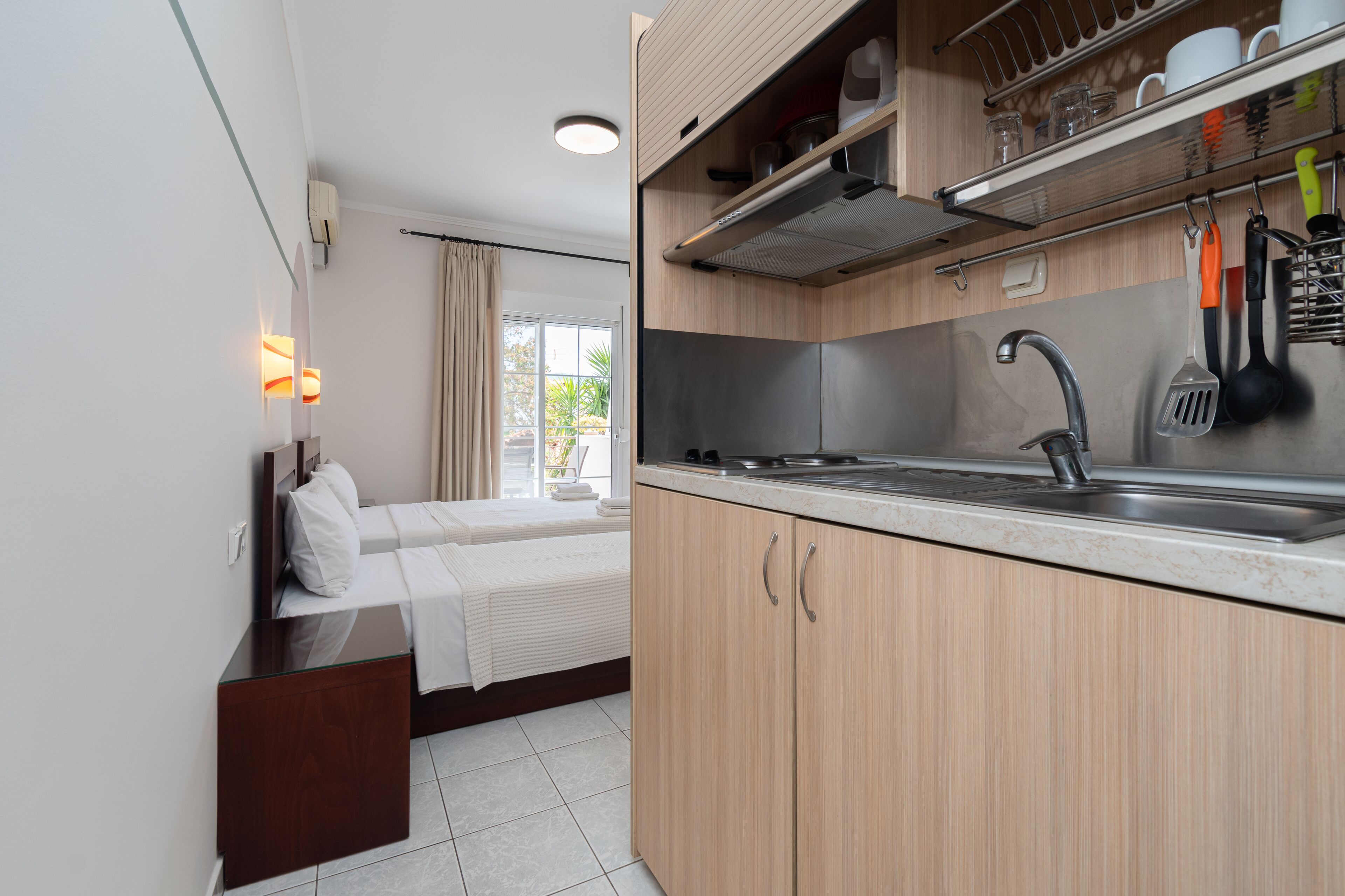 Quarto de hotel compacto com cozinha compacta, uma cama com roupa de cama branca, mesa de cabeceira e janela com cortinas.