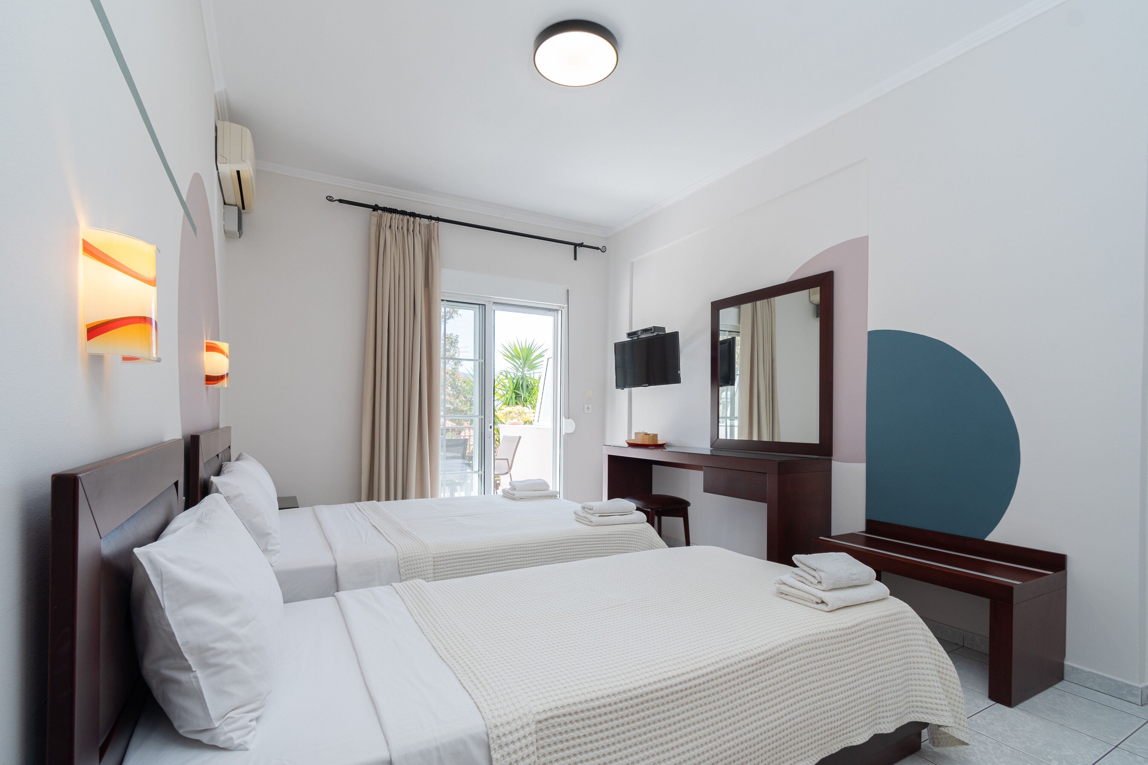 Quarto de hotel com duas camas, uma escrivaninha, uma cadeira, uma televisão e uma janela grande com cortinas.