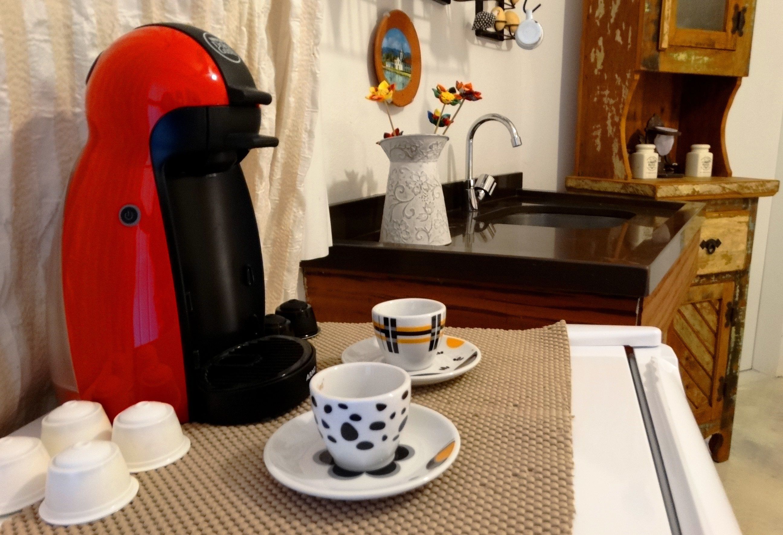 Uma cozinha compacta com cafeteira, duas xícaras, um vaso com flores e uma pia.