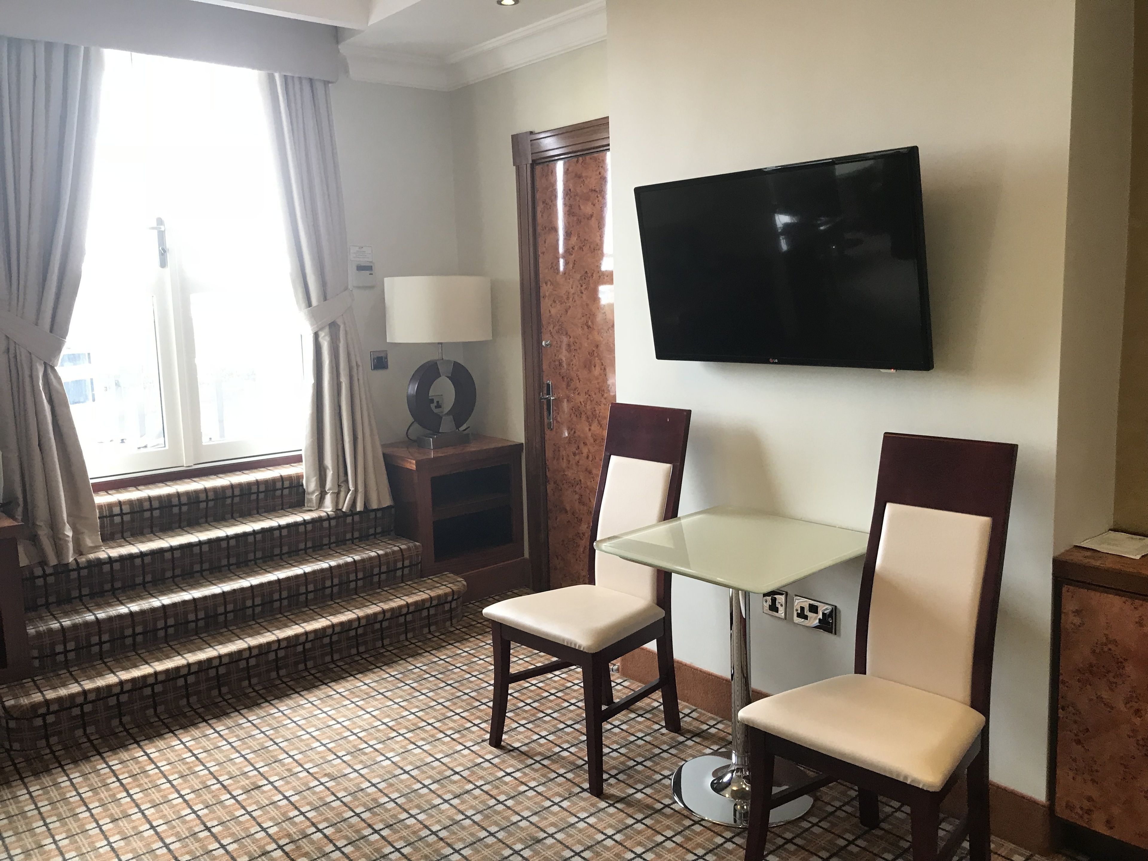 Quarto de hotel com televisão na parede, uma mesa pequena com abajur, duas cadeiras, uma janela com cortinas e um chão estampado.