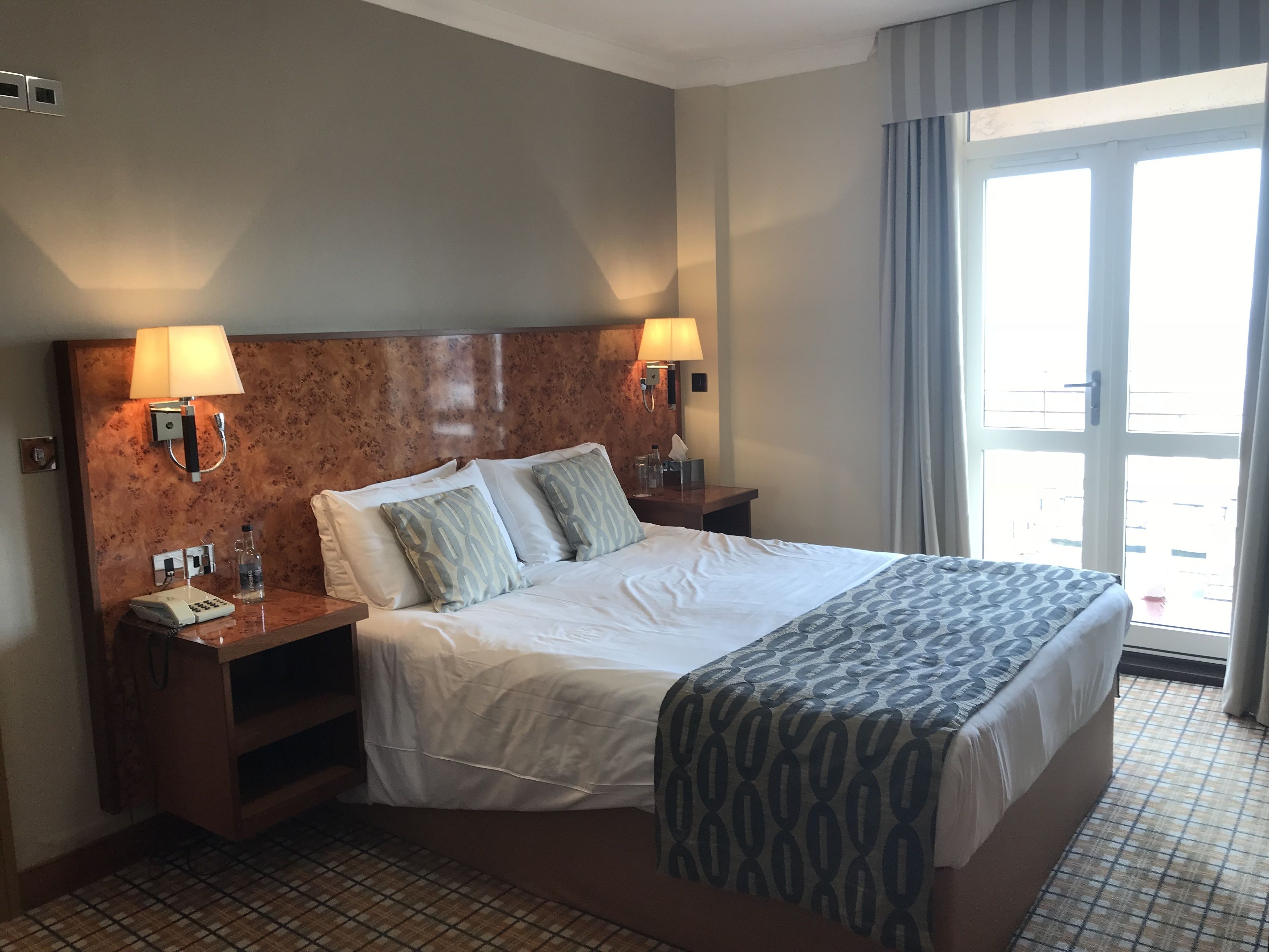 Quarto de hotel com uma cama grande, duas luminárias de cabeceira, um telefone, uma mesinha e uma janela com cortinas.