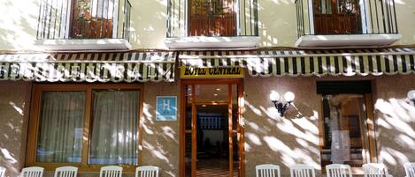 Entrada de hotel com uma placa indicando 'HOTEL CENTRAL', um conjunto de cadeiras de plástico branco na frente e um chão de azulejos.