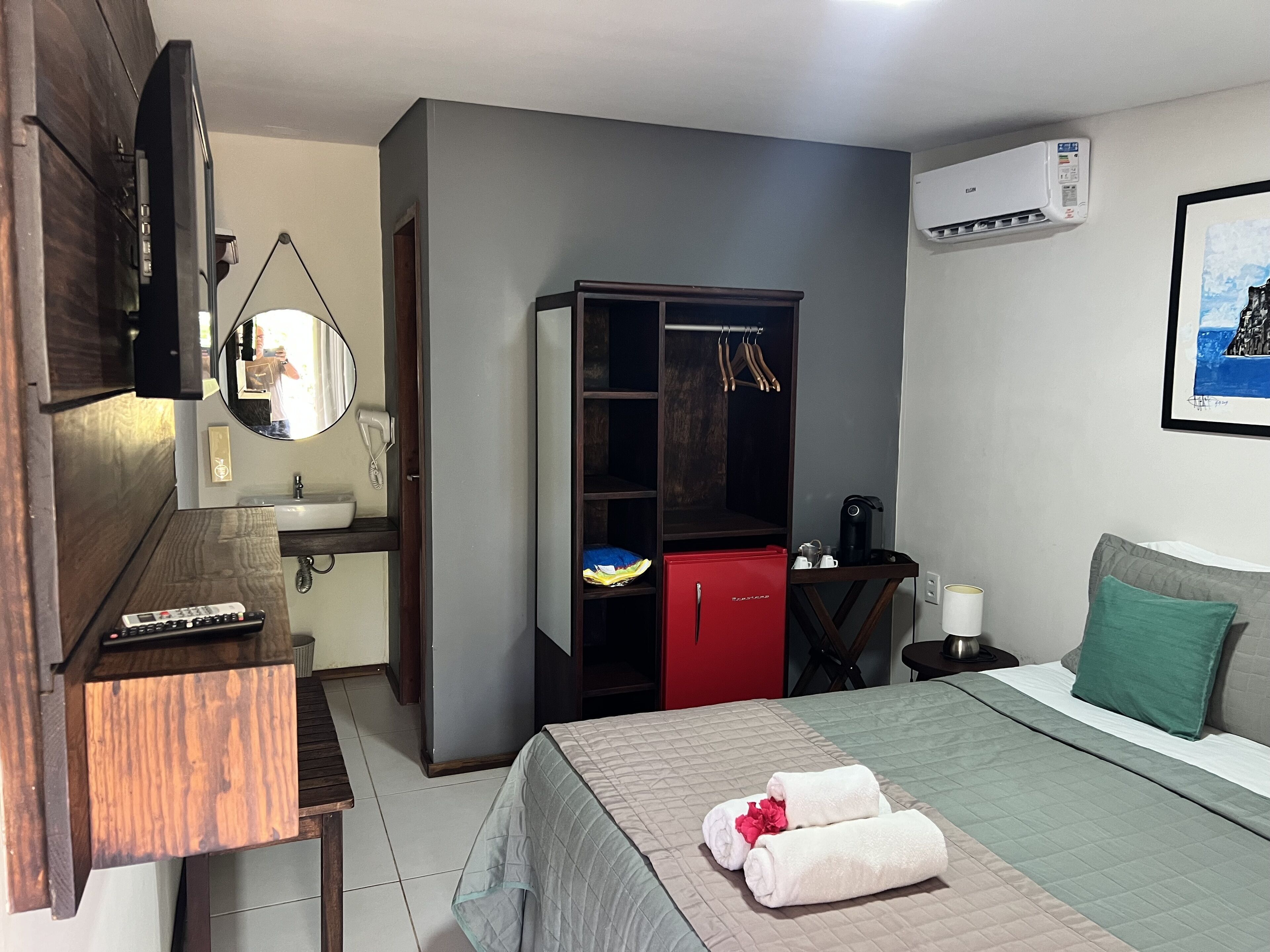 Quarto de hotel com cama, guarda-roupa, secretária com espelho, pia e ar condicionado.