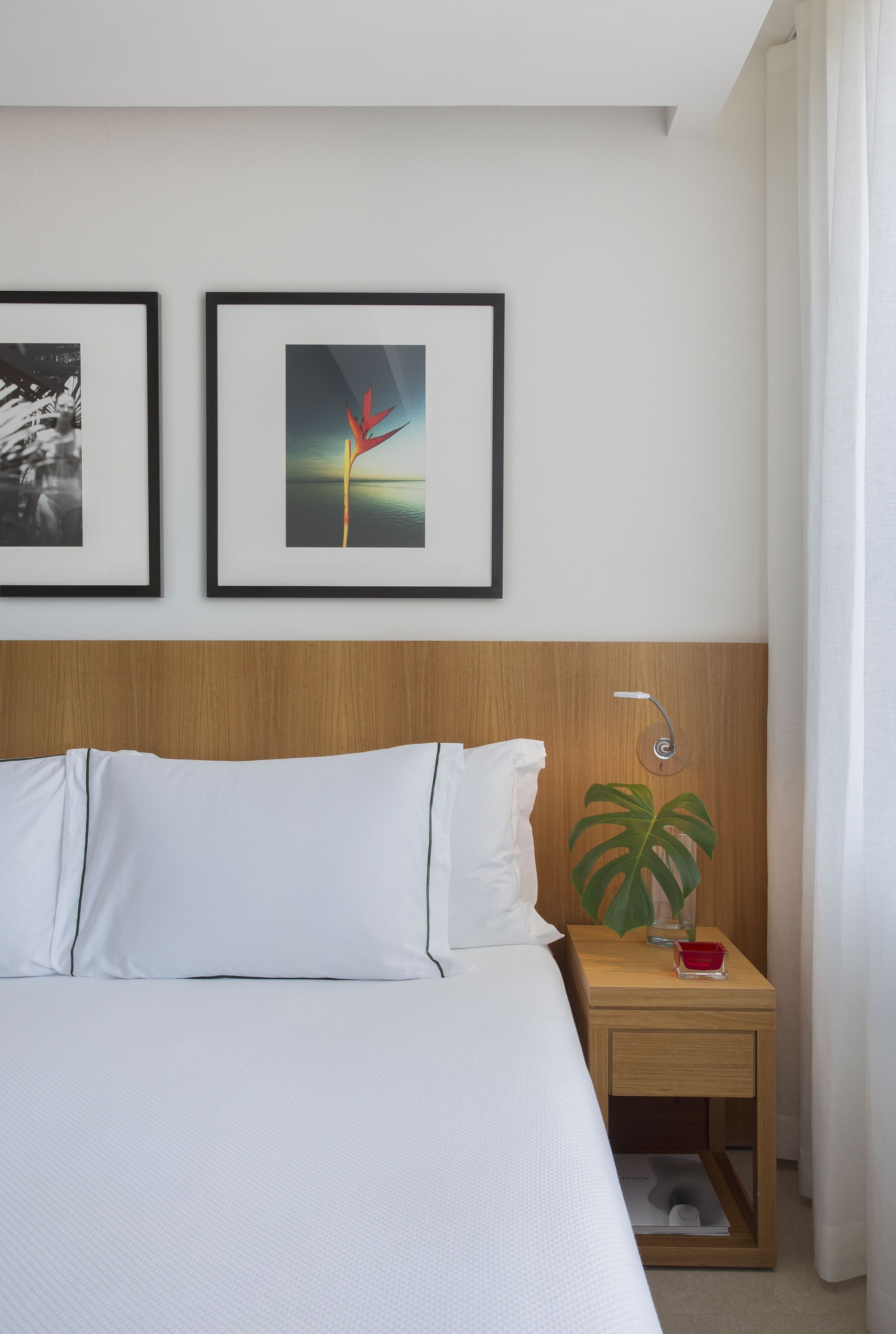 Quarto de hotel com cama, mesa de cabeceira de madeira, um quadro emoldurado e um vaso com planta.