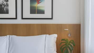 Quarto de hotel com cama, mesa de cabeceira de madeira, um quadro emoldurado e um vaso com planta.