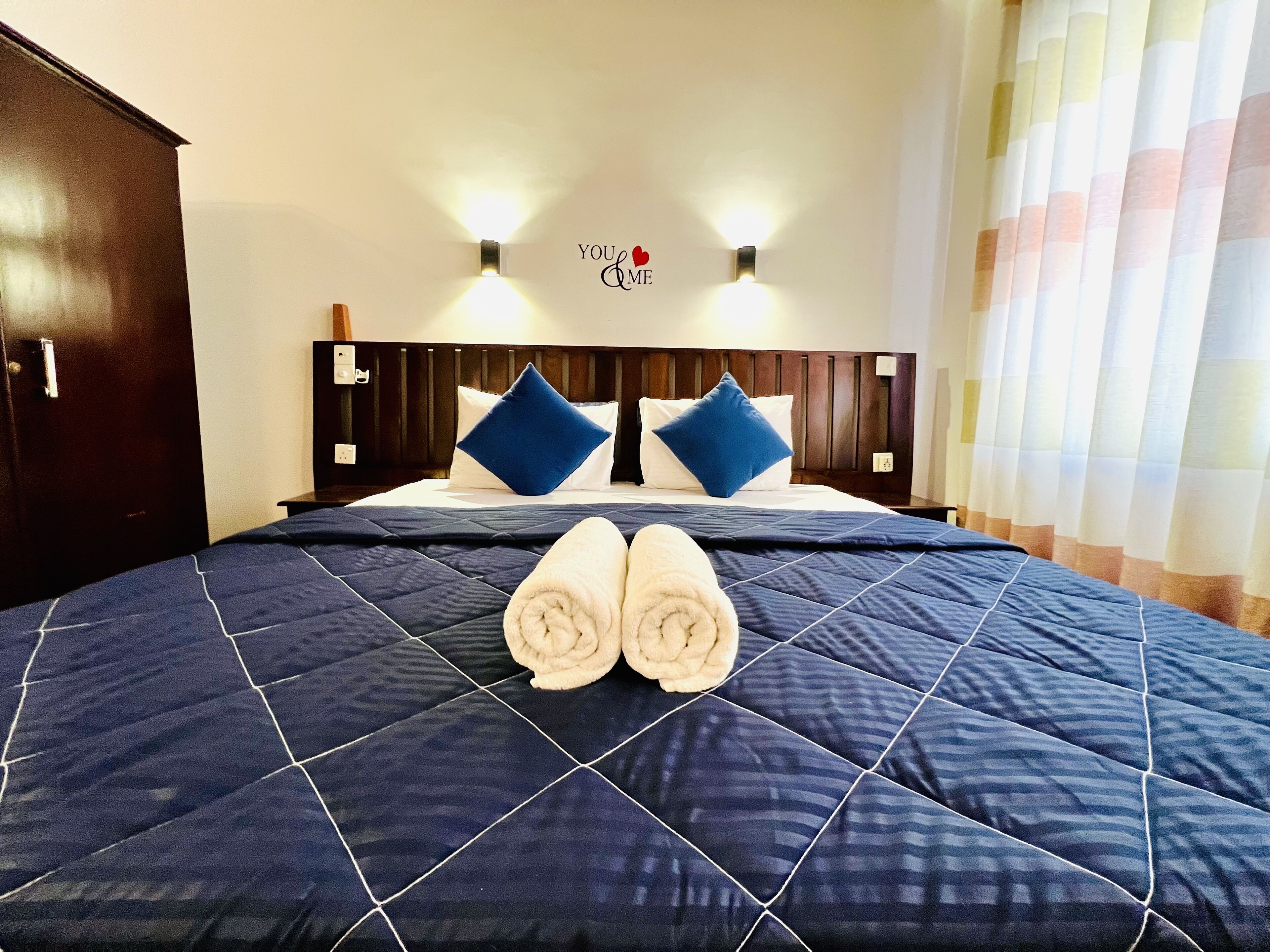 Quarto de hotel com cama, duas luzes fixadas na parede, um letreiro com a inscrição 'You & Me' e janela com cortinas.