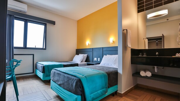 Quarto de hotel com duas camas, uma escrivaninha, uma cadeira, uma janela e banheiro com espelho e pia.