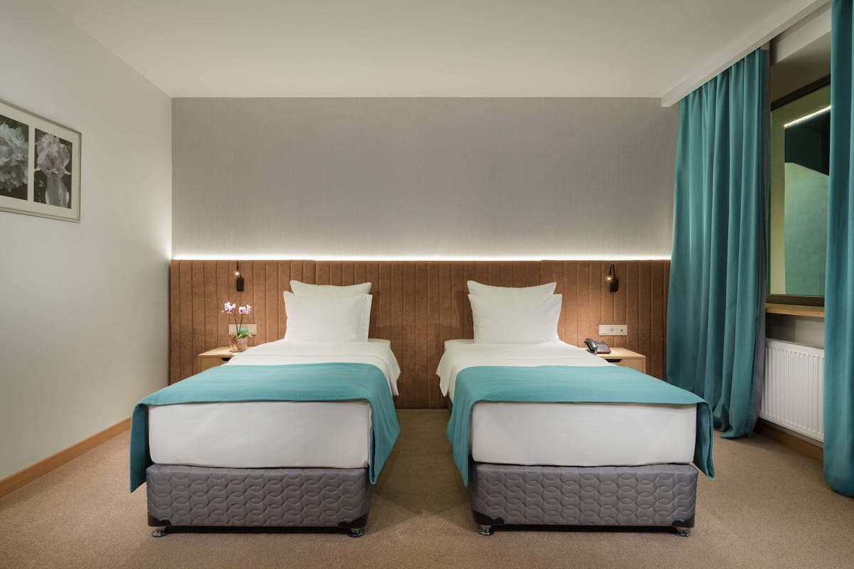 Quarto de hotel com duas camas, cabeceira de madeira, mesa de cabeceira com abajur e janela com cortinas azul-turquesa.