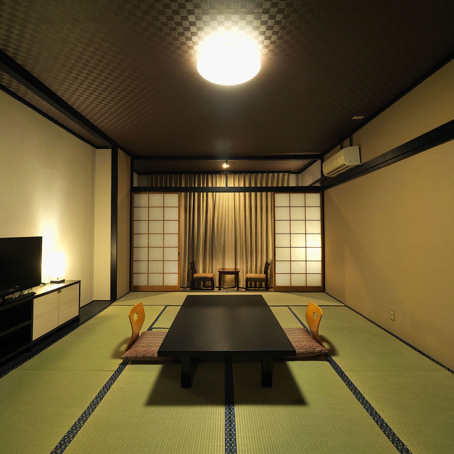 Um quarto em estilo japonês tradicional, com piso de tatame, uma mesa baixa, cadeiras, uma televisão e portas corrediças.