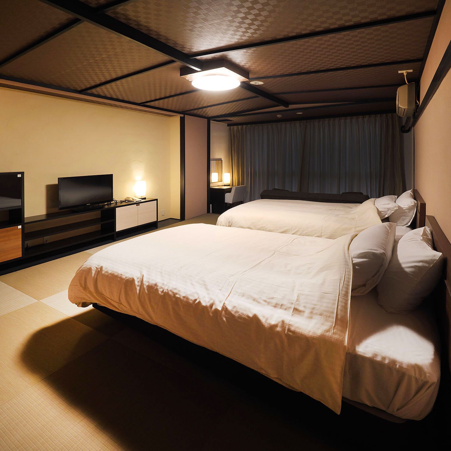 Quarto de hotel com cama, televisão, escrivaninha com luminária e iluminação no teto.