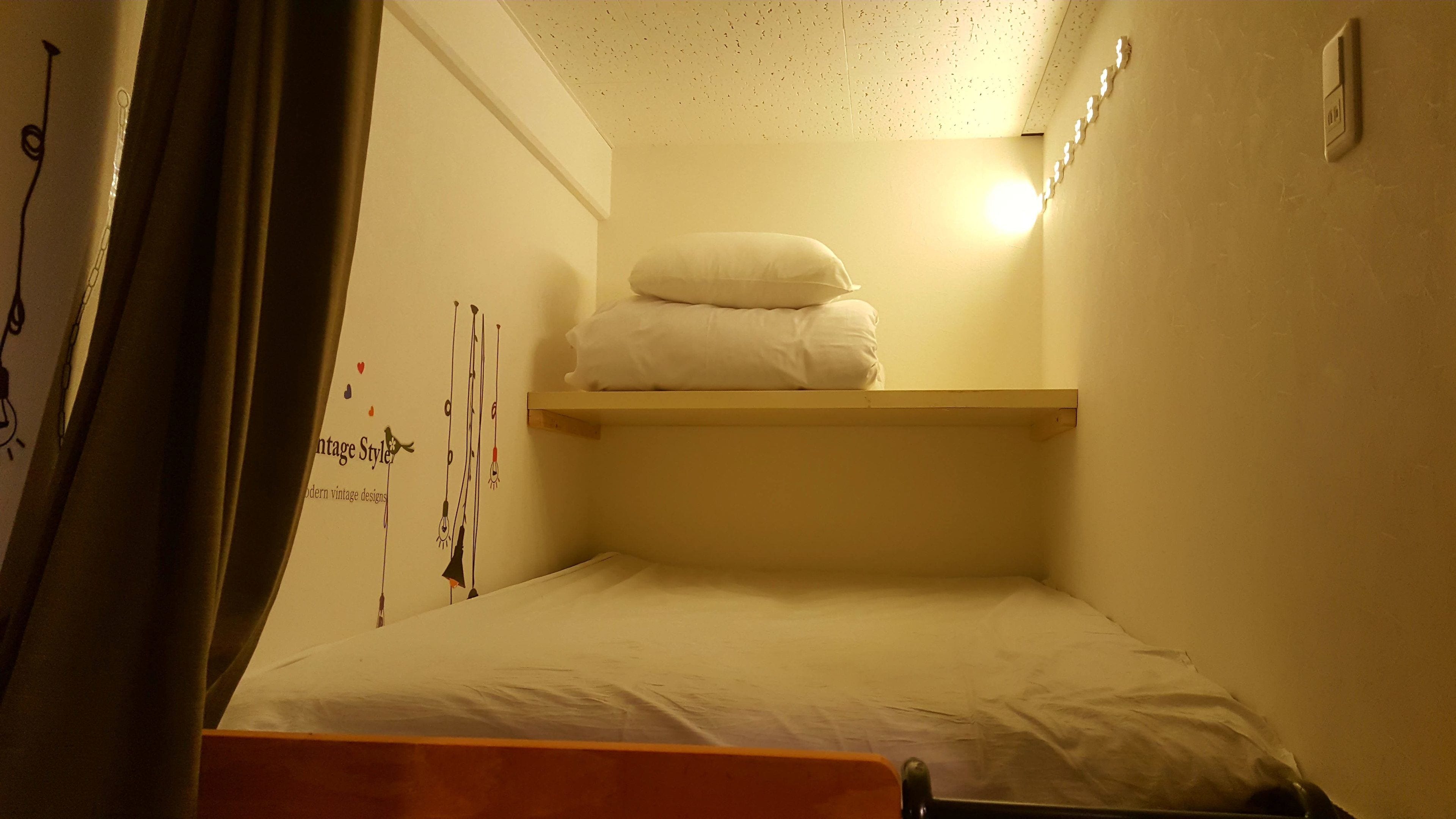 Quarto pequeno e estreito com uma cama de solteiro, um prateleira sobre a cama e um painel com interruptor de luz e tomada.