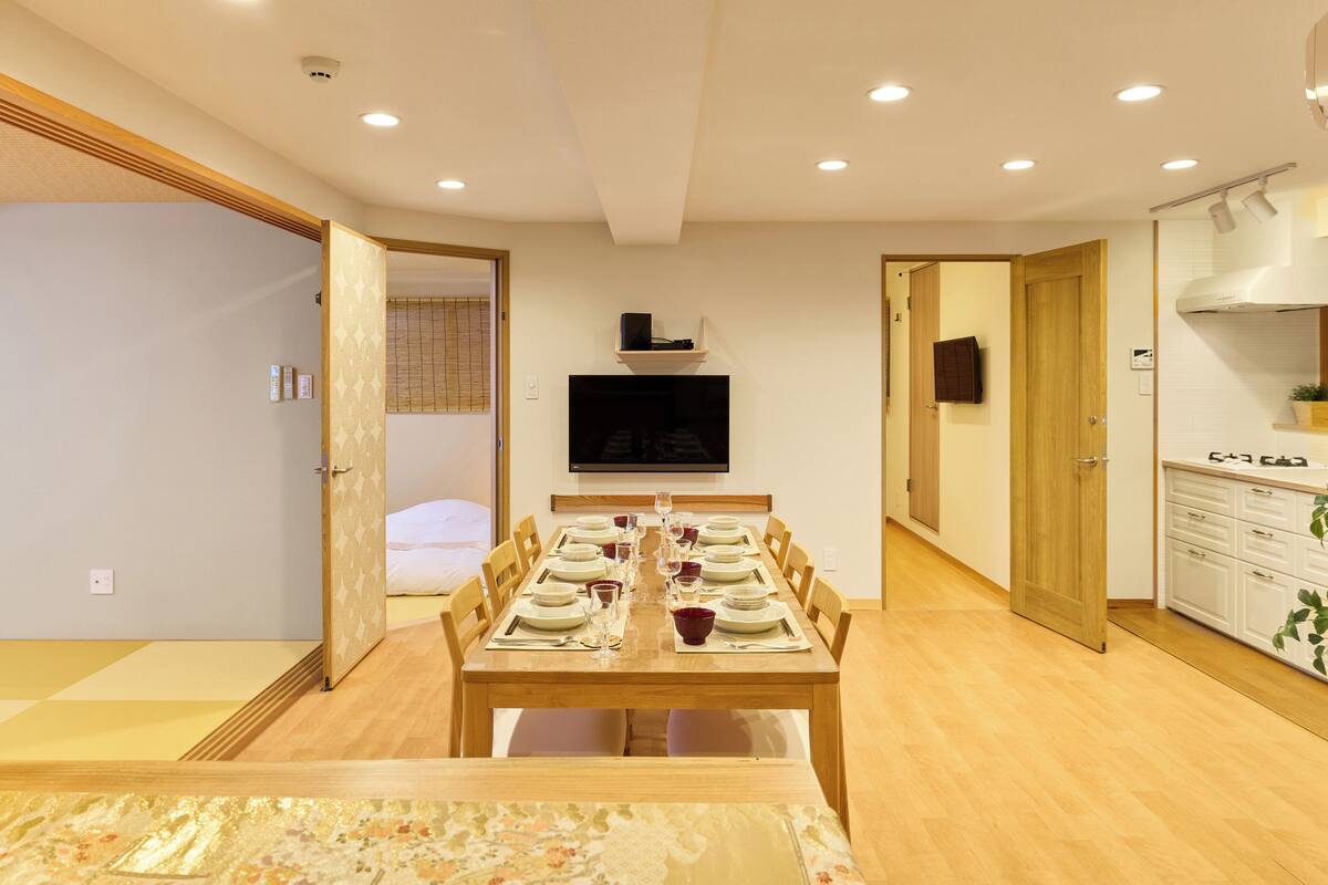 Uma área de refeições com uma mesa de madeira posta para quatro pessoas, uma televisão fixada na parede e uma cozinha com armários e fogão.