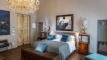 Quarto com cama grande, lustre, pintura, baú de madeira e um banco.
