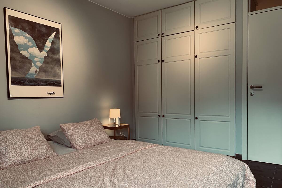 Quarto com cama, mesa de cabeceira, abajur, guarda-roupa e um quadro na parede.