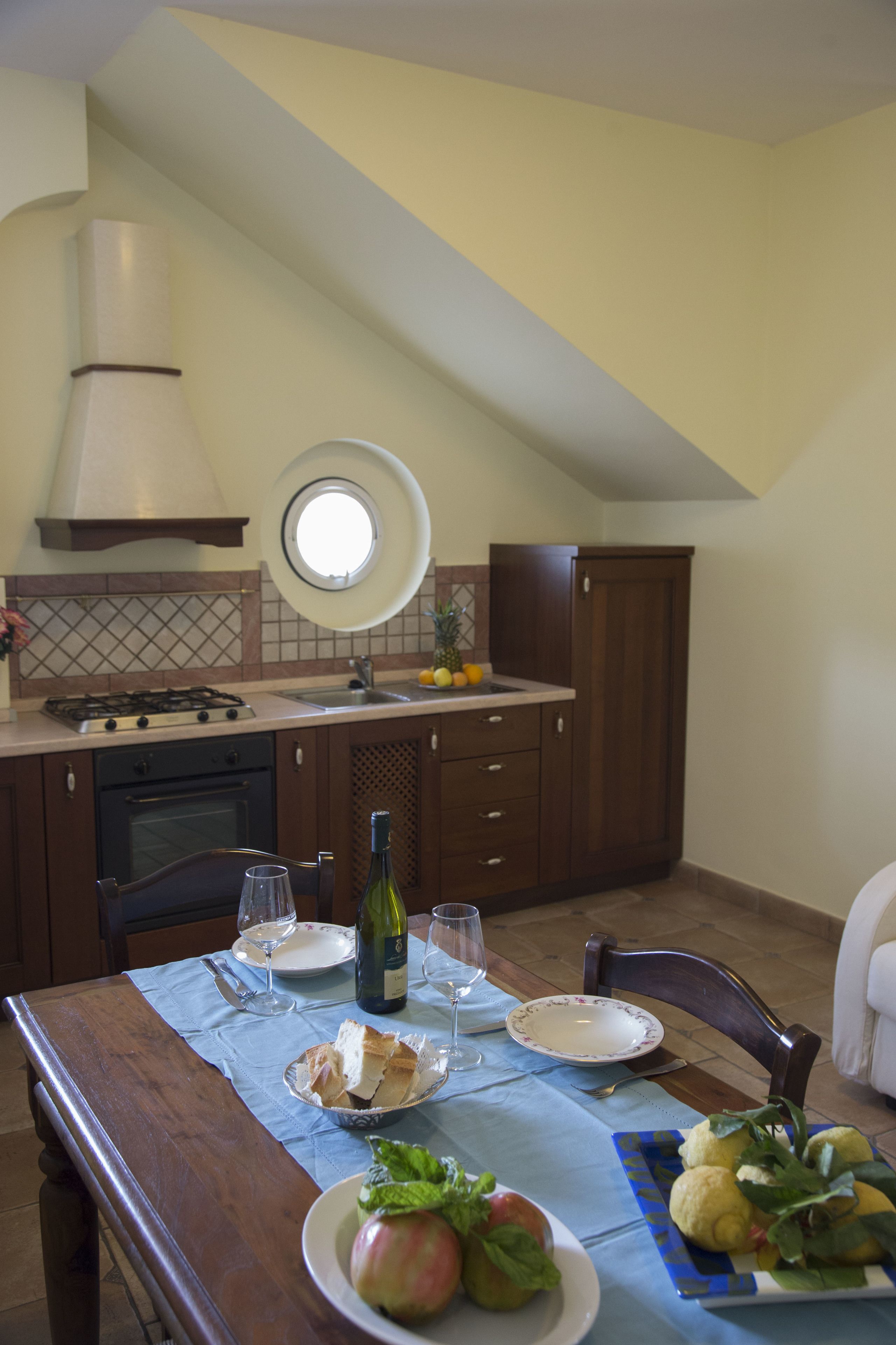 Uma área de refeições com uma mesa posta para dois, com uma travessa de frutas, pão e uma garrafa de vinho.