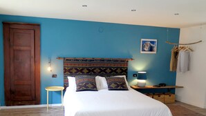 Quarto com cama, porta de madeira, parede azul, luminária suspensa, mesa pequena e prateleira fixada na parede.