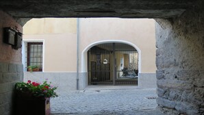 Um pátio de calçada com muro de pedra, um vaso de planta e um portão metálico que dá acesso a um edifício com fachada bege e entrada em arco.