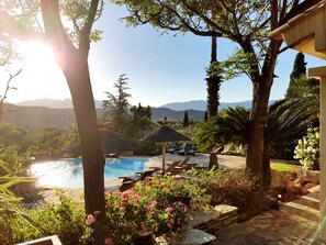 Área de piscina com espreguiçadeiras, uma estrutura com telhado de palha e vista para montanhas.
