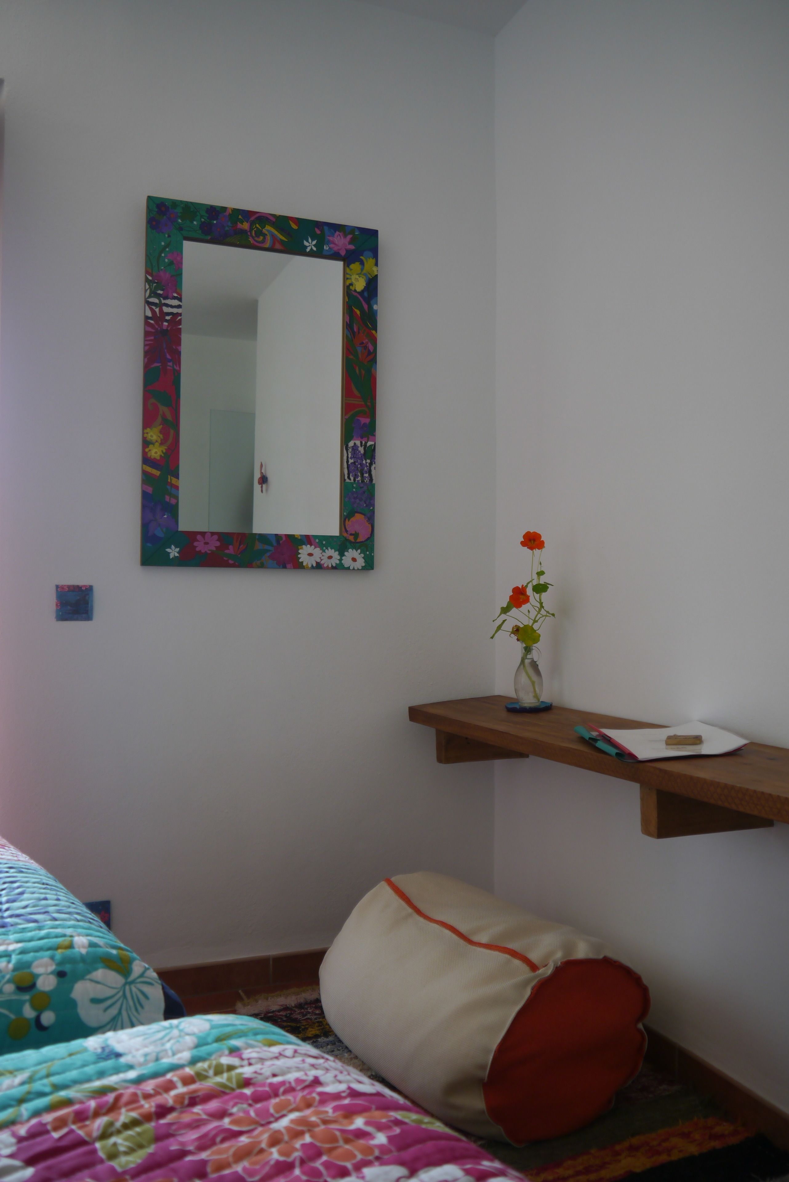 Um quarto com um espelho colorido, um pequeno vaso com flores, uma prateleira de madeira e uma cama com colcha colorida.