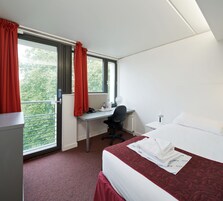 Quarto de hotel com cama, escrivaninha, cadeira, radiador e janela com cortinas.