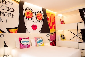 Quarto de hotel moderno com um pôster grande de uma mulher usando óculos e texto chamativo que diz "NEED PRACTICE MORE". Há beliches com almofadas e uma luminária na mesinha de cabeceira.