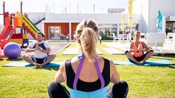 Um grupo de pessoas meditando sobre tapetes de yoga em área externa, com um parquinho e um edifício ao fundo.