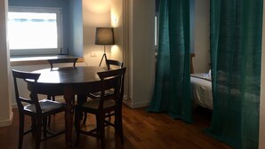Um quarto com uma mesa de jantar e cadeiras, uma luminária, uma cama com cortina verde e uma janela com persianas.