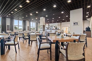 Uma sala de jantar espaçosa com várias mesas e cadeiras, uma cozinha ao fundo e grandes janelas que permitem entrada de luz natural.