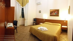 Quarto de hotel com cama, escrivaninha, cadeira, janela com cortinas, quadro na parede e ar condicionado.