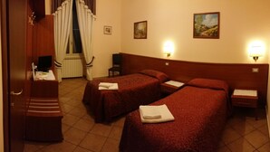 Quarto de hotel com duas camas, um guarda-roupa de madeira, uma escrivaninha com cadeira, uma janela com cortinas e luminárias fixadas na parede.