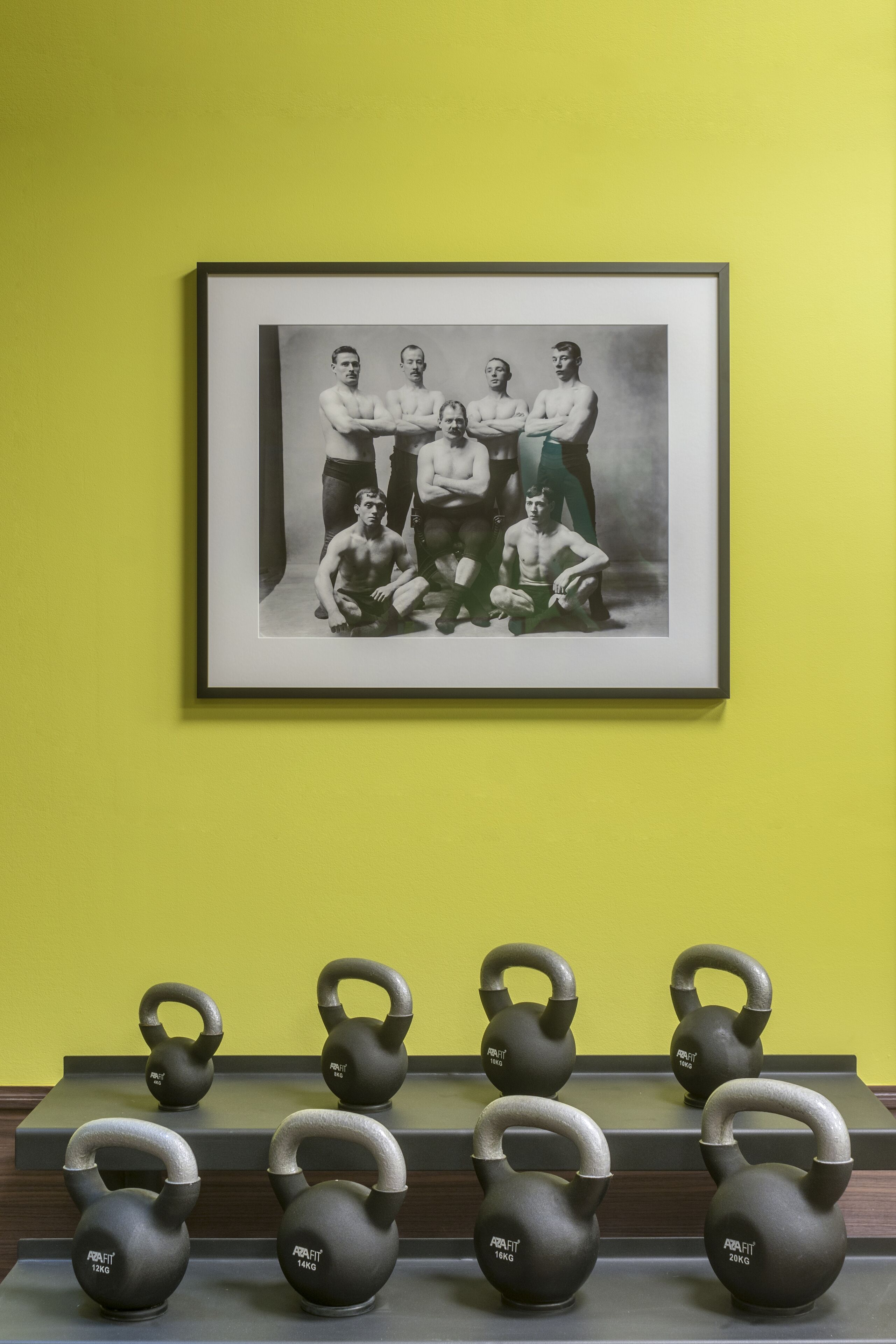 Fotografia a preto e branco emoldurada numa parede amarela, com halteres em baixo.