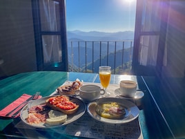 Mesa com itens de café da manhã, um copo de suco de laranja e uma xícara de café, com vista para montanhas através de uma varanda.