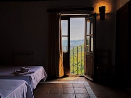 Quarto com cama, mesa e toalhas, e vista para o verde através de uma porta aberta.